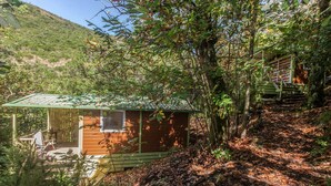 Uma cabana de madeira com telhado verde, rodeada por folhagem densa e uma encosta arborizada.