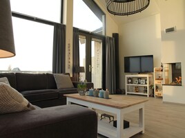 Uma sala de estar moderna com um sofá preto, uma mesa de centro de madeira e uma televisão sobre um suporte branco.