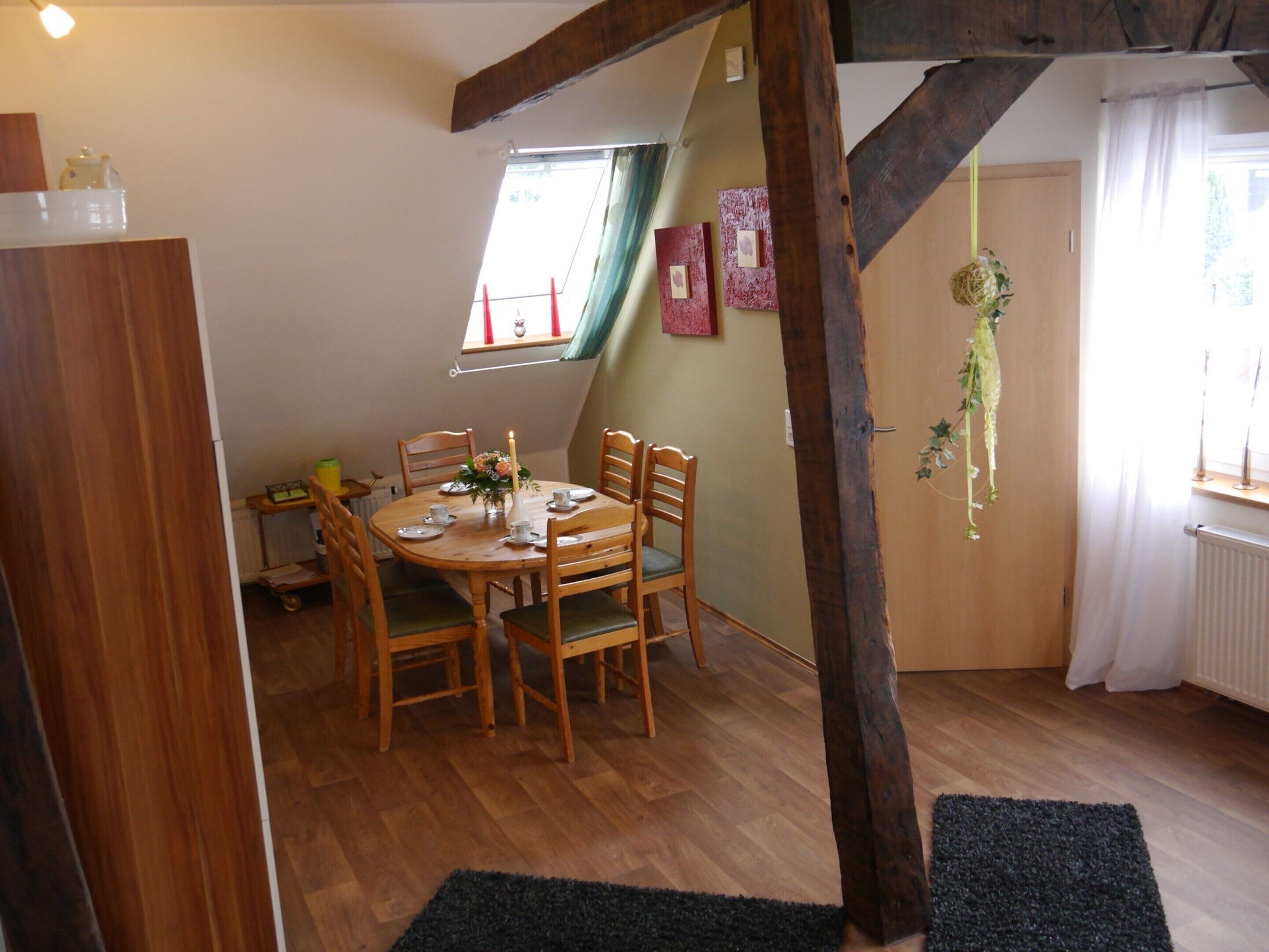Uma área de refeições com mesa e cadeiras de madeira, janela com cortinas e uma planta pendurada.