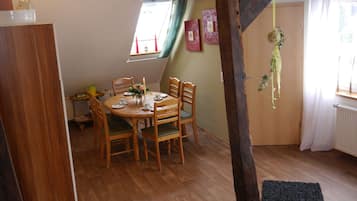 Uma área de refeições com mesa e cadeiras de madeira, janela com cortinas e uma planta pendurada.