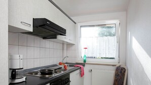 Cozinha compacta com armários brancos, fogão preto, cafeteira e janela com vista para uma cerca e árvores.