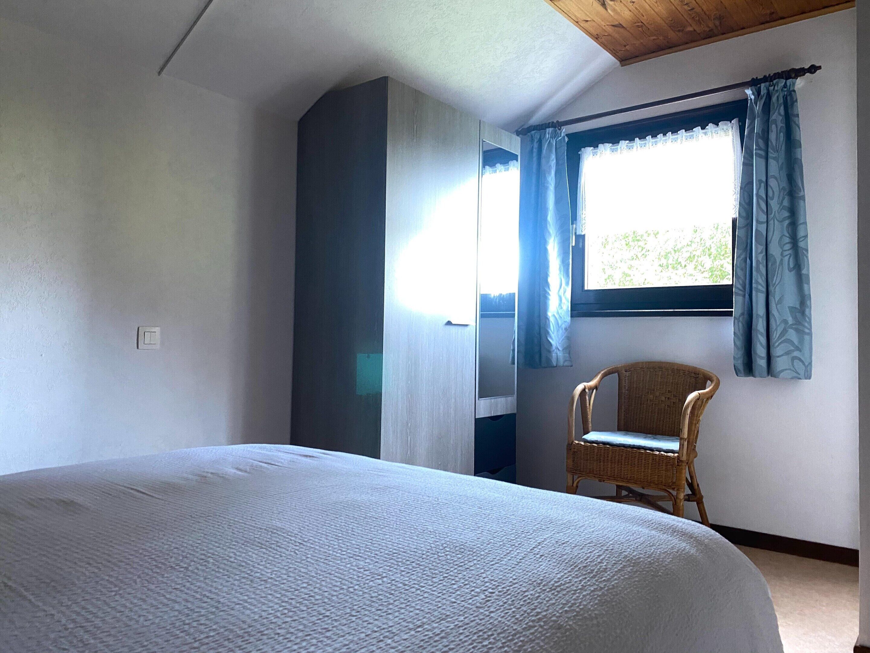 Quarto com cama grande, uma cadeira de vime, janela com cortinas e teto de madeira.