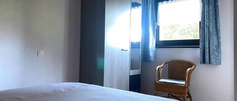 Quarto com cama grande, uma cadeira de vime, janela com cortinas e teto de madeira.