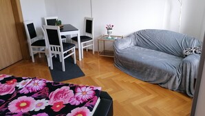 Quarto com sofá cinzento, uma mesa pequena com uma planta, cadeiras brancas e uma cama com colcha estampada.