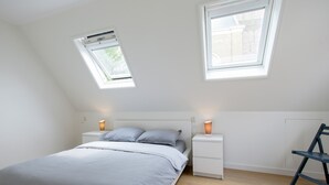 Quarto com cama, duas mesas de cabeceira, escrivaninha e duas janelas.