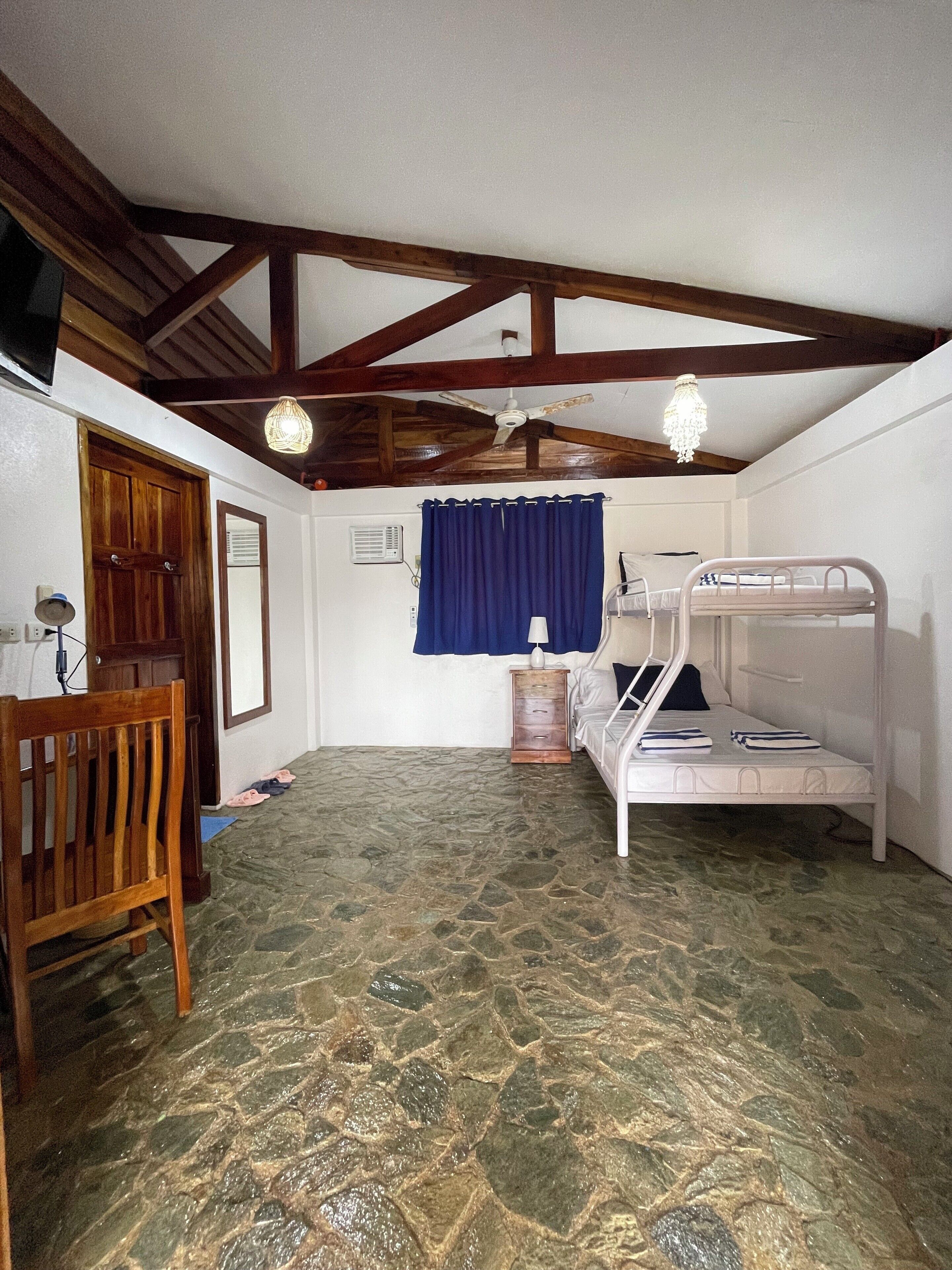 Quarto com cama de solteiro e beliche, uma cadeira de madeira, escrivaninha, espelho e janela com cortinas azuis.
