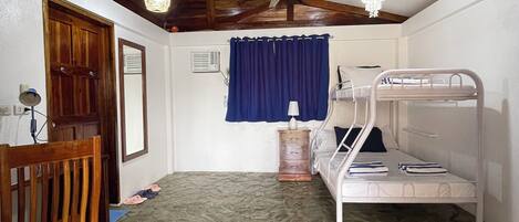 Quarto com cama de solteiro e beliche, uma cadeira de madeira, escrivaninha, espelho e janela com cortinas azuis.