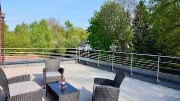 Terraço no telhado com mobiliário de vime e uma mesa, com vista para um jardim e um edifício.