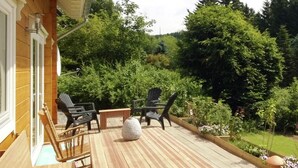 Um deck de madeira com mobiliário de exterior, um vaso de pedra e um jardim com diversas plantas e árvores.