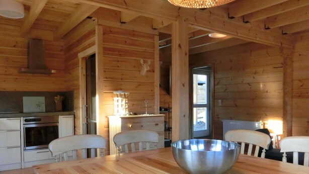 Interior em madeira com mesa de jantar, cadeiras, uma tigela de metal grande, cozinha com eletrodomésticos e uma janela.