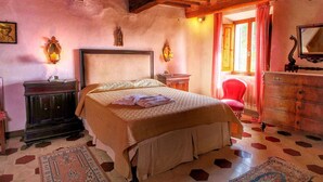 Quarto com uma cama grande, cabeceira de madeira e uma cadeira vermelha. Há um aparador com um espelho e uma pequena mesa com uma luminária. O quarto possui piso de azulejo e uma janela com cortinas vermelhas.
