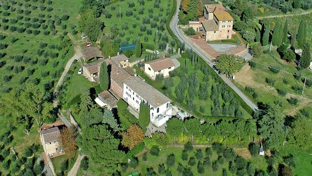 Vista aérea de uma propriedade rural com vários edifícios, incluindo uma casa grande e branca com telhado de telhas, cercada por vegetação exuberante e um pequeno lago.