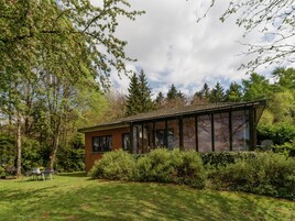 Uma casa moderna com uma estufa de vidro, rodeada por vegetação exuberante e árvores.