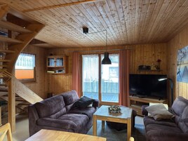 Interior de cabana de madeira com sofá, mesinha, televisão, escada e janela com cortinas.