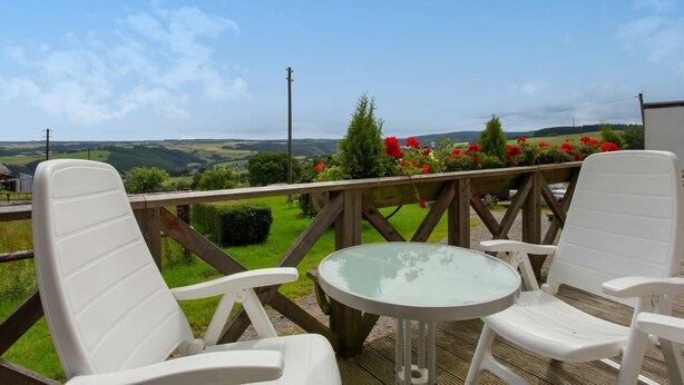 Um terraço com cadeiras de plástico branco e uma mesa de vidro, com vista para uma paisagem deslumbrante com flores vermelhas e campos verdes.