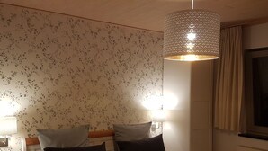 Quarto com cama, duas almofadas, mesa de cabeceira, abajur e parede com papel de parede floral.