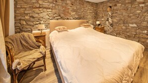 Quarto com parede de pedra, cama com roupa de cama branca, mesa de cabeceira de madeira com abajur e cadeira de vime com manta.