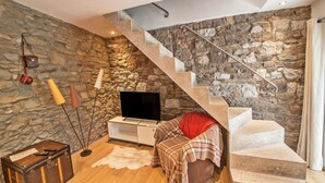 Uma sala de estar aconchegante com parede de pedra, televisão de tela plana, poltrona de couro, mala antiga e escada.