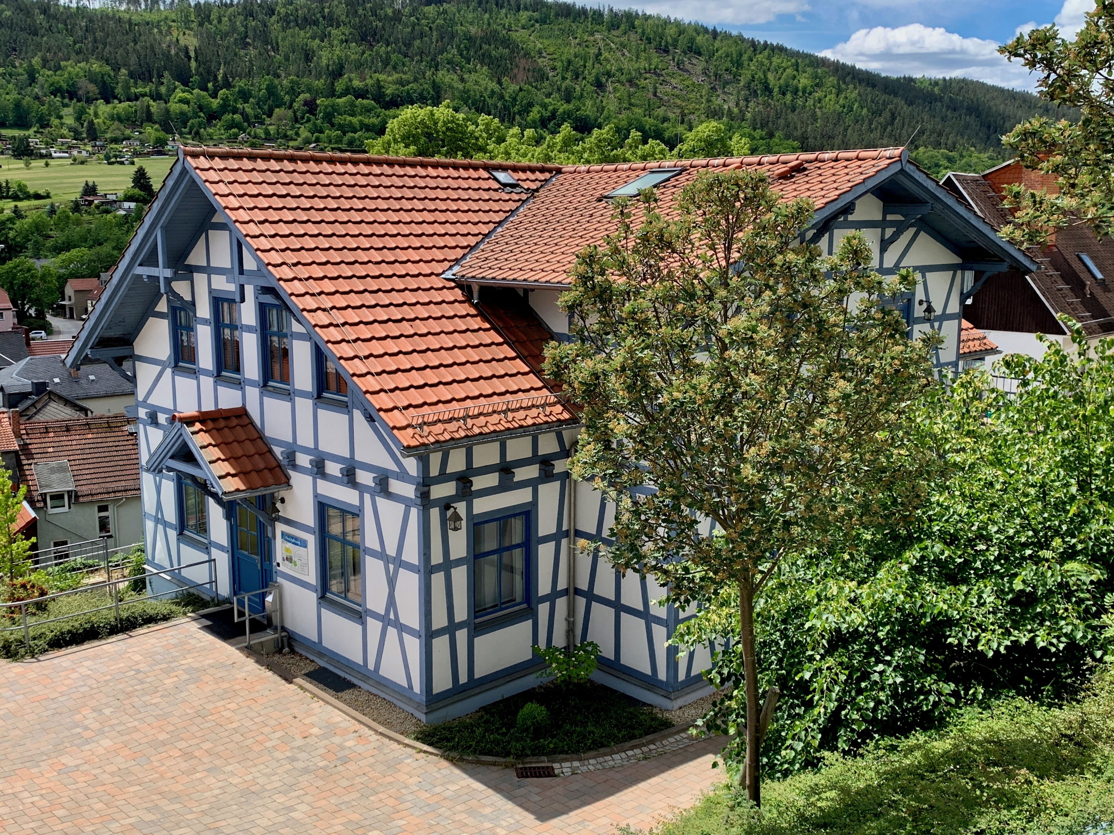 Uma casa tradicional em estilo de enxaimel, com telhado de telhas vermelhas, rodeada por vegetação exuberante e uma encosta arborizada.