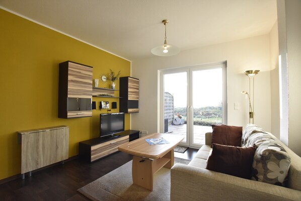 Uma sala de estar moderna com sofá, mesa de centro e televisão. Há uma janela com vista para o exterior.