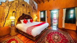 Quarto com cama grande, cabeceira de madeira e um mural de um edifício na parede. Há duas portas e uma janela com cortinas.