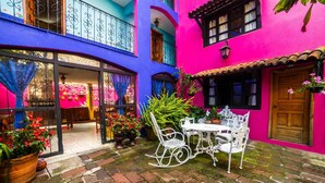 Um pátio com uma cadeira de balanço branca e uma mesa, cercado por edifícios coloridos com varandas e plantas em vasos.