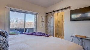 Quarto moderno com uma cama grande, um guarda-roupa com porta de celeiro deslizante, uma TV na parede e uma janela com vista para árvores.