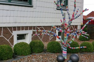 Uma casa com fachada branca, uma parede de pedra marrom e uma escultura de árvore colorida feita de tricô no jardim da frente.