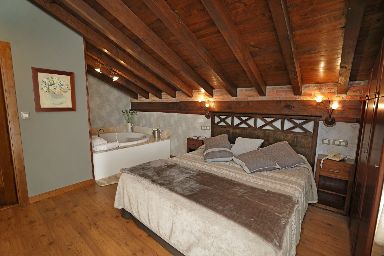 Quarto com teto de madeira, uma cama grande com várias almofadas, mesa de cabeceira, luminária fixada na parede e banheiro com pia e banheira.
