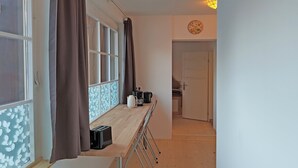 Corredor estreito com uma mesa de madeira, chaleira e torradeira. Uma porta dá acesso a uma cozinha.