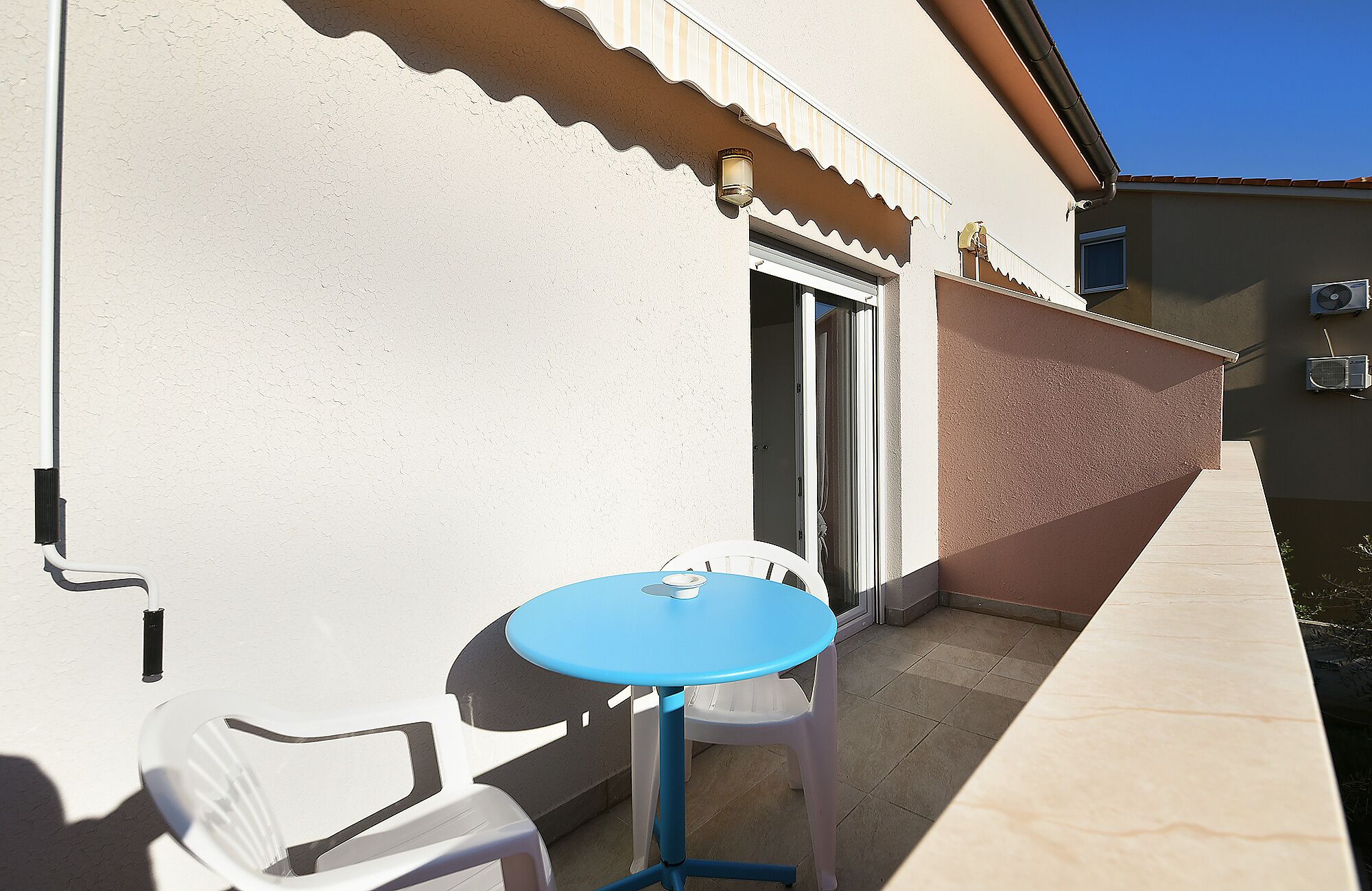 Uma pequena área de estar ao ar livre com uma mesa azul e cadeiras brancas, próxima a um edifício de fachada branca e parede com detalhes em marrom.
