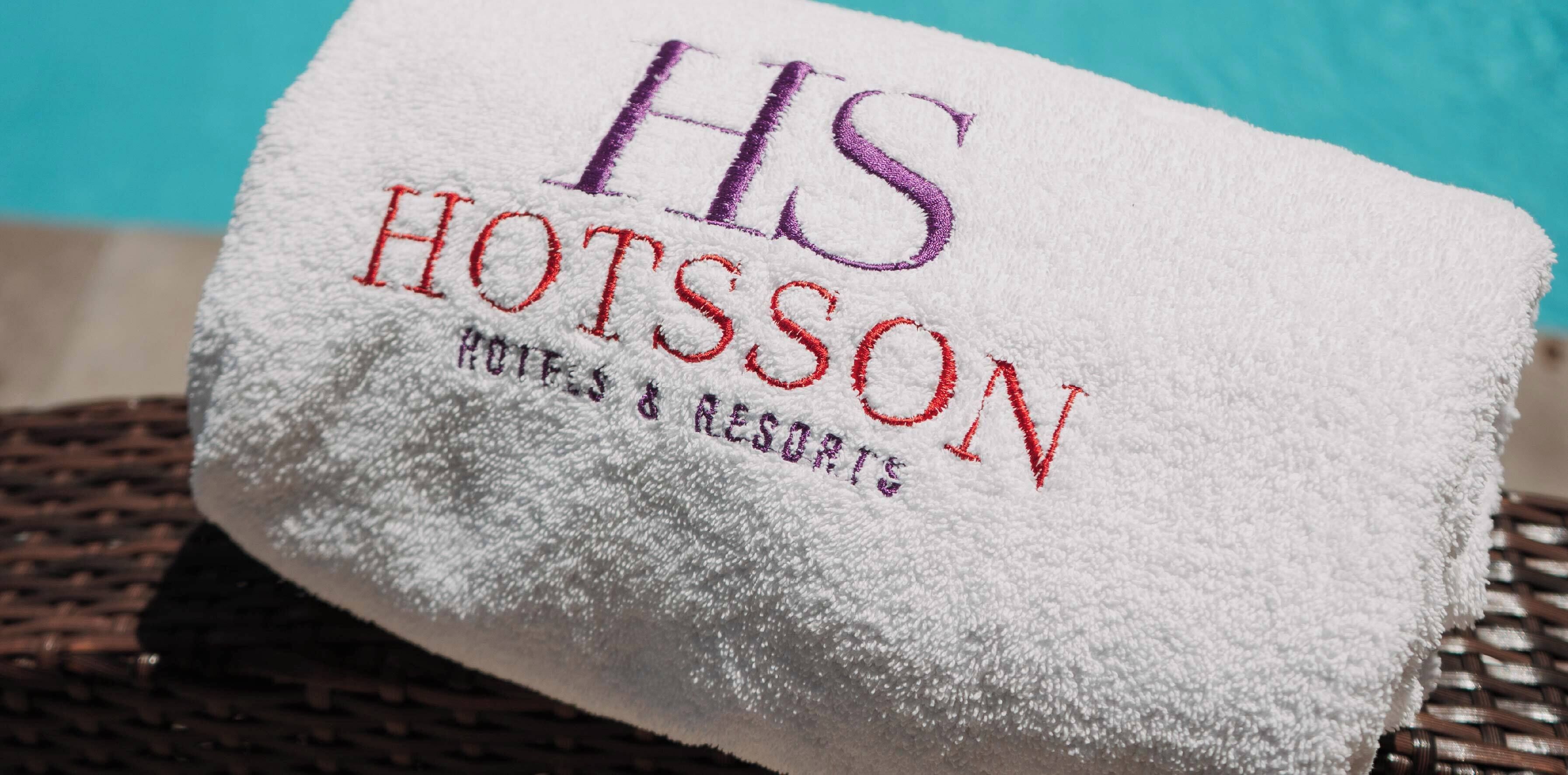 Toalha branca com bordado contendo o texto "HS HOTSSON ROYAL & RESORTS".