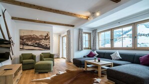 Uma sala de estar espaçosa com um teto de madeira grande, um tapete de couro, um sofá, uma mesa de centro e uma pintura de uma paisagem nevada.