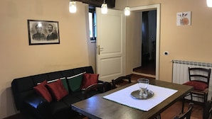 Uma sala de jantar com mesa de madeira, cadeiras e um sofá preto com almofadas vermelhas. Há uma fotografia emoldurada na parede e um pequeno vaso com flores.