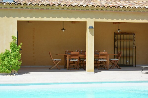 Uma área de refeições coberta ao ar livre, com piscina, cadeiras de madeira e uma mesa.