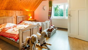 Um quarto aconchegante com teto inclinado, paredes de madeira, uma cama com cobertor xadrez vermelho e branco, uma mesa de cabeceira com abajur, uma janela com vista para o verde e uma porta.