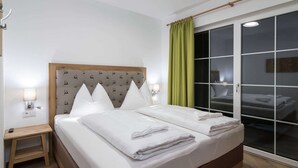 Quarto de hotel com cama, abajures, mesa de cabeceira, porta de vidro deslizante e piso de madeira.