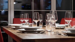 Uma mesa de jantar posta com vários copos de vinho, pratos e talheres, com cadeiras estofadas em vermelho e uma janela ao fundo.