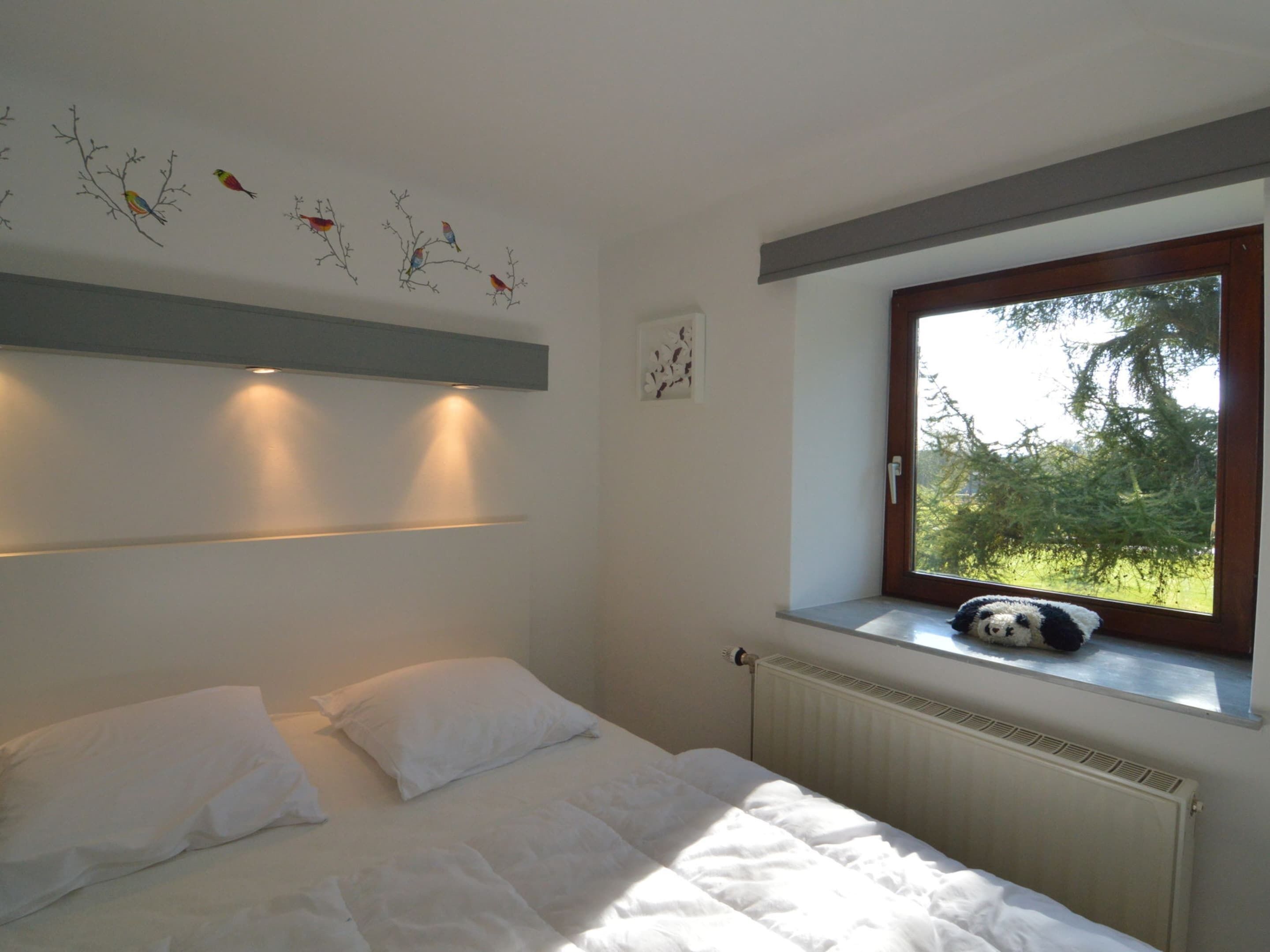 Quarto com cama, almofadas brancas, janela com vista para árvores e arte nas paredes com pássaros.