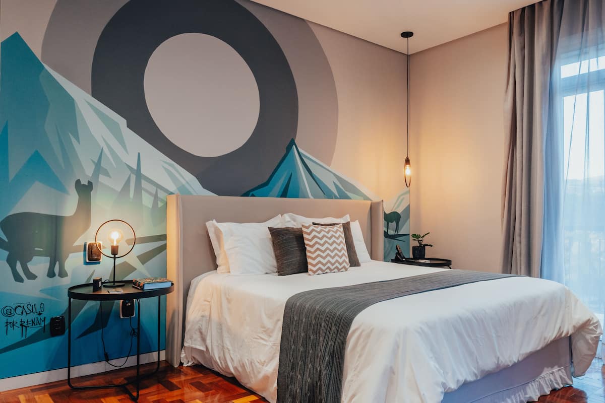 Quarto de hotel moderno com uma cama grande, uma mesa de cabeceira, um mural na parede e uma janela com cortinas.