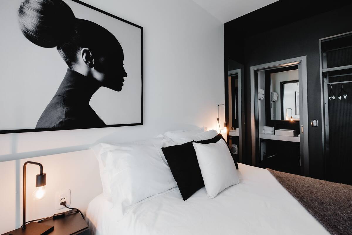 Quarto de hotel com cama, mesa de cabeceira, luminária na parede, retrato emoldurado e porta de acesso a um banheiro.