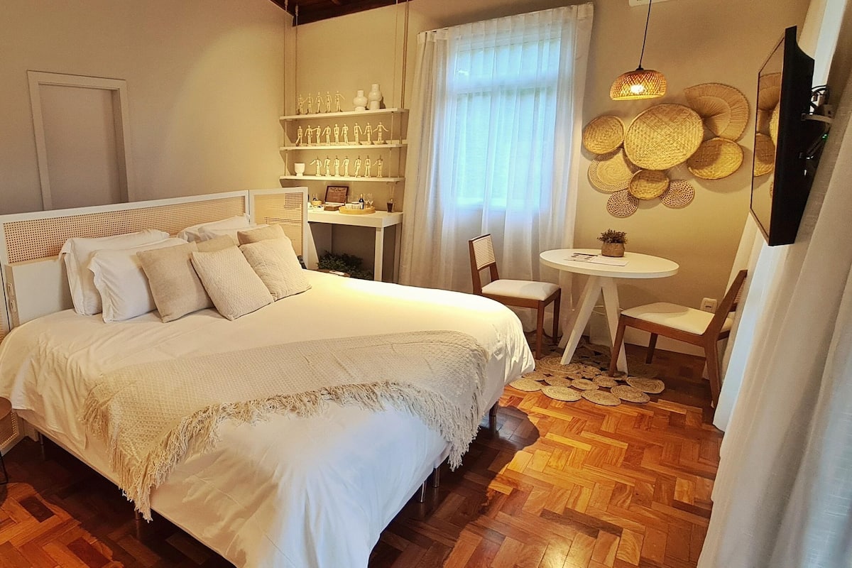 Quarto com uma cama grande, uma mesinha, uma cadeira e uma estante com objetos decorativos.