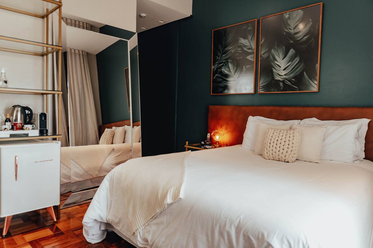 Quarto de hotel com uma cama grande, mesa de cabeceira com abajur, guarda-roupa e secretária com máquina de café.