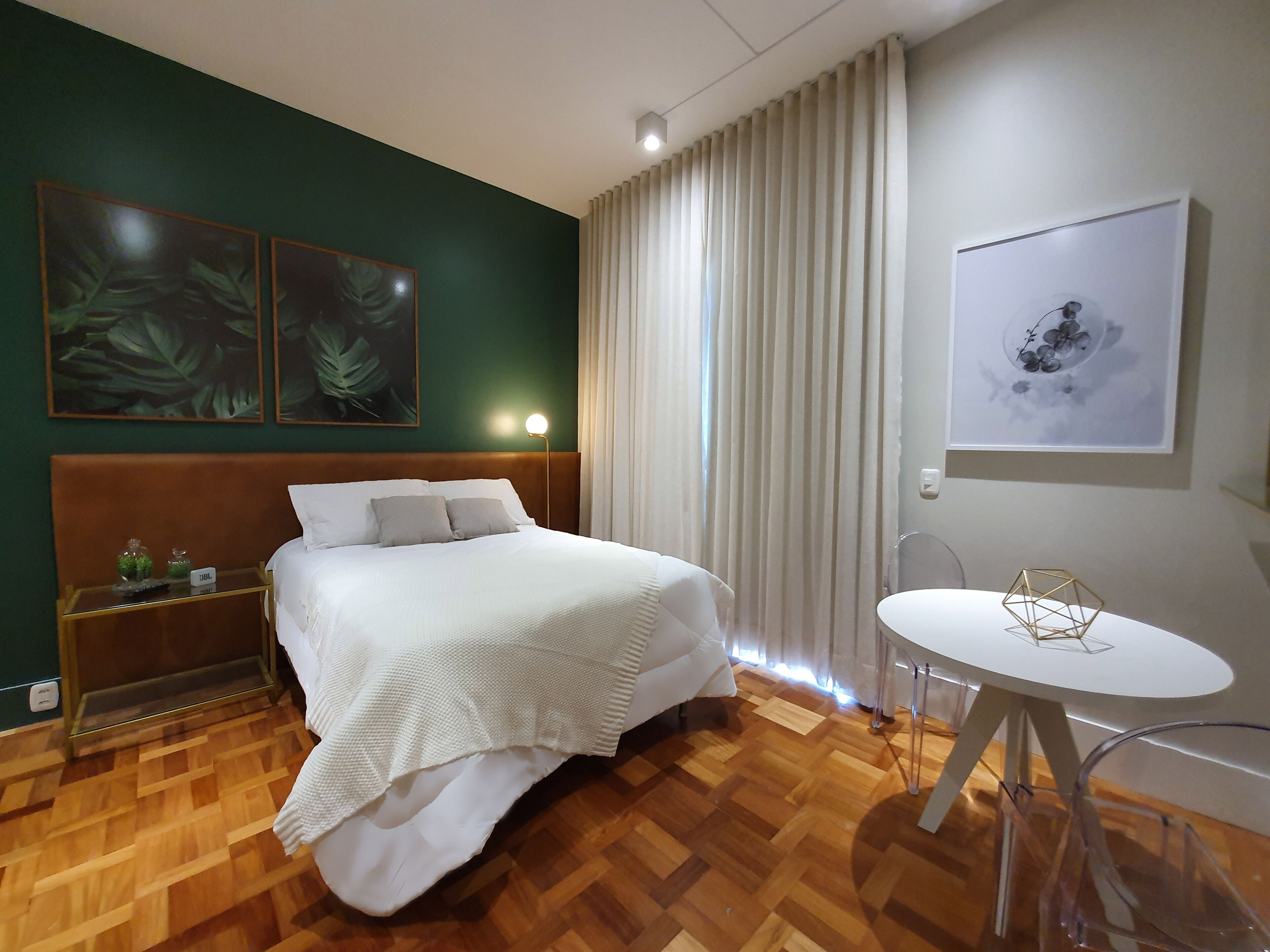 Quarto de hotel com cama, mesa de cabeceira com abajur, mesa redonda com cadeiras e obras de arte nas paredes.