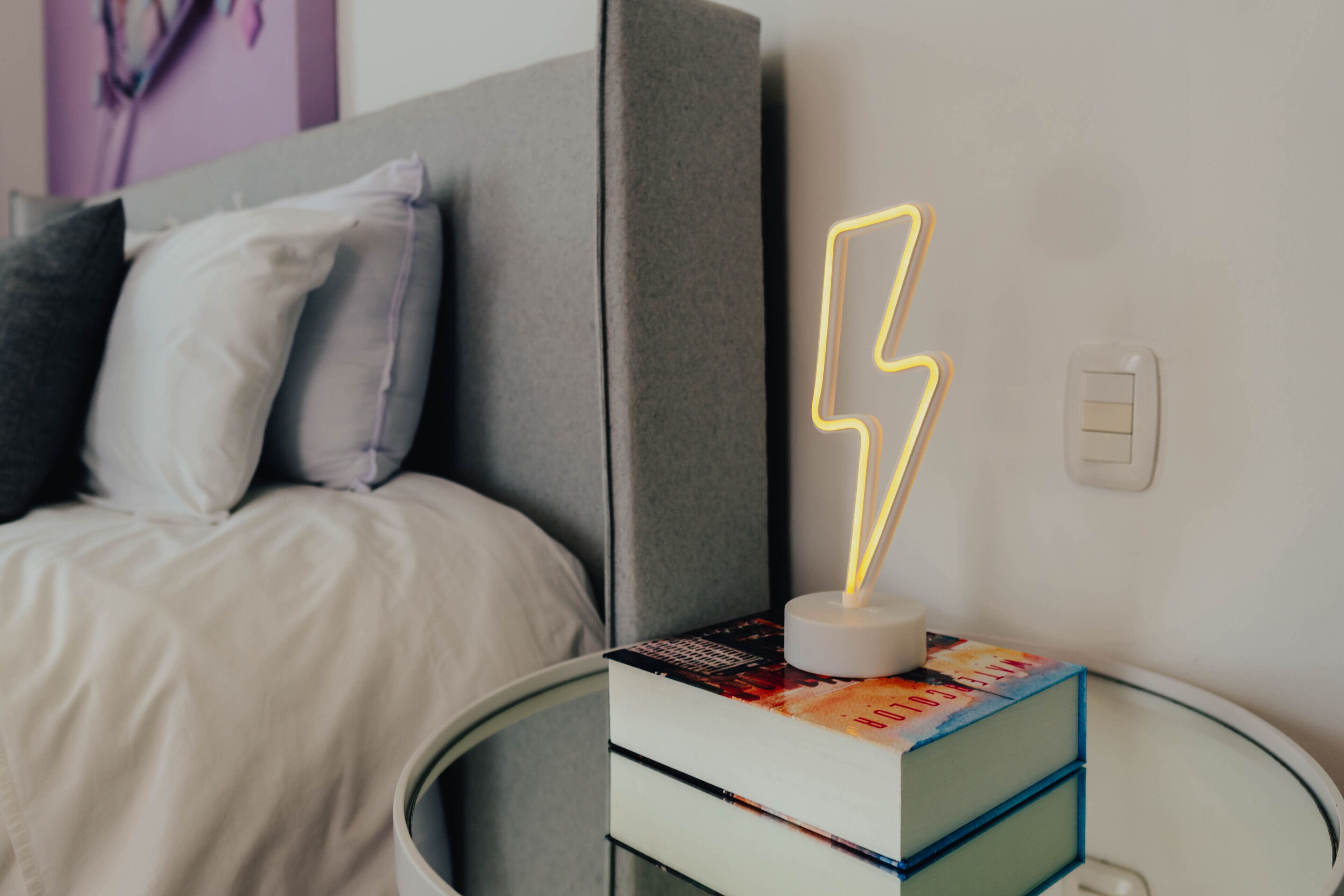 Cama bem arrumada com almofadas, mesa de cabeceira com um livro e uma luminária decorativa, e um interruptor na parede.