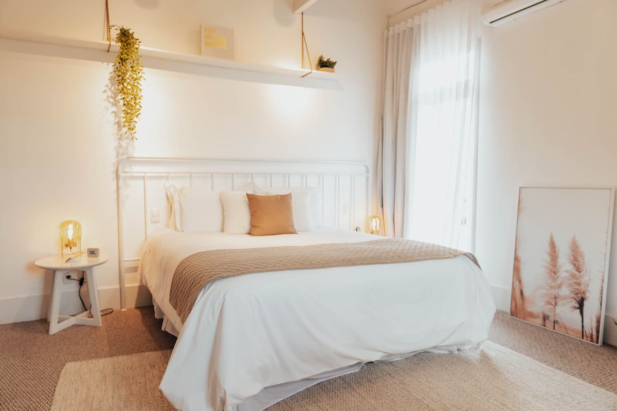 Quarto com cama branca, mesa de cabeceira com abajur, planta pendente e quadro na parede.