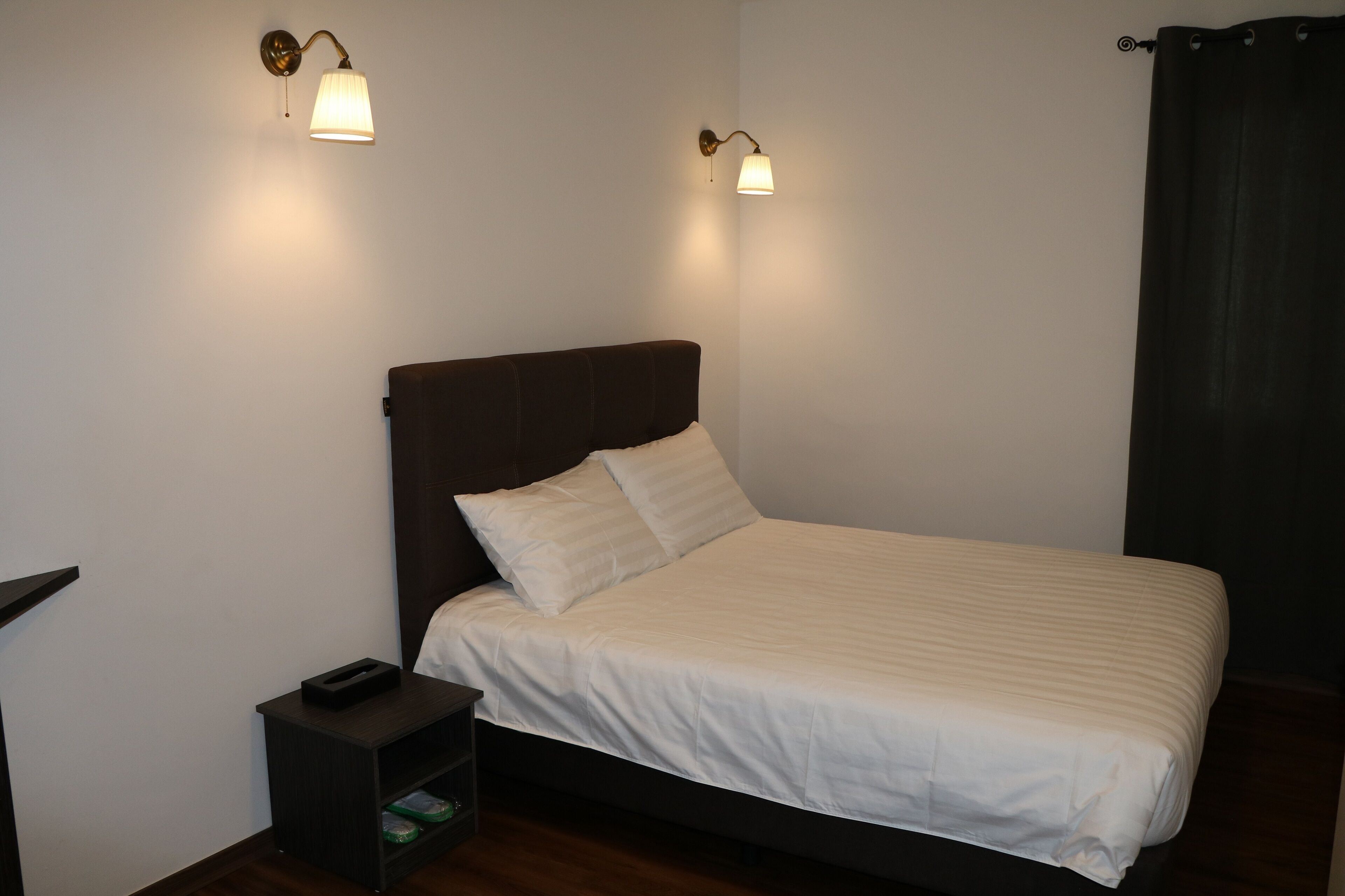 Cama bem arrumada com roupa de cama branca, cabeceira escura e duas luminárias fixadas na parede.