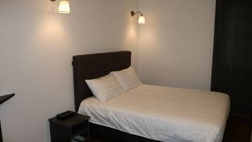 Cama bem arrumada com roupa de cama branca, cabeceira escura e duas luminárias fixadas na parede.