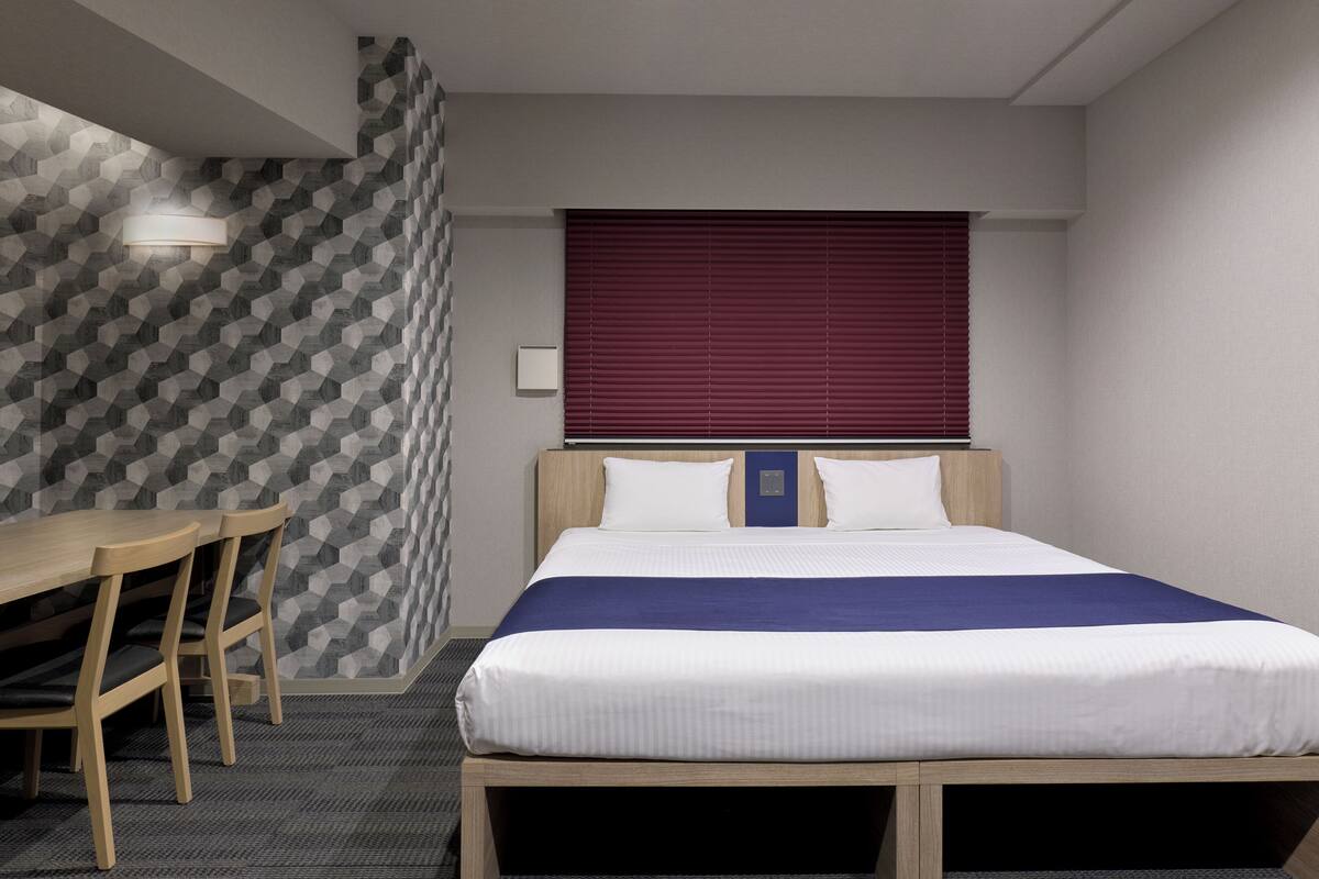 Quarto de hotel com cama, escrivaninha, duas cadeiras, janela com persianas e parede estampada.