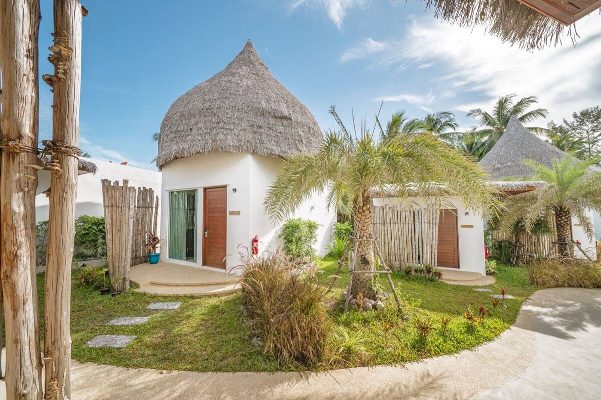 Uma cabana com telhado de palha, rodeada por vegetação tropical e palmeiras.